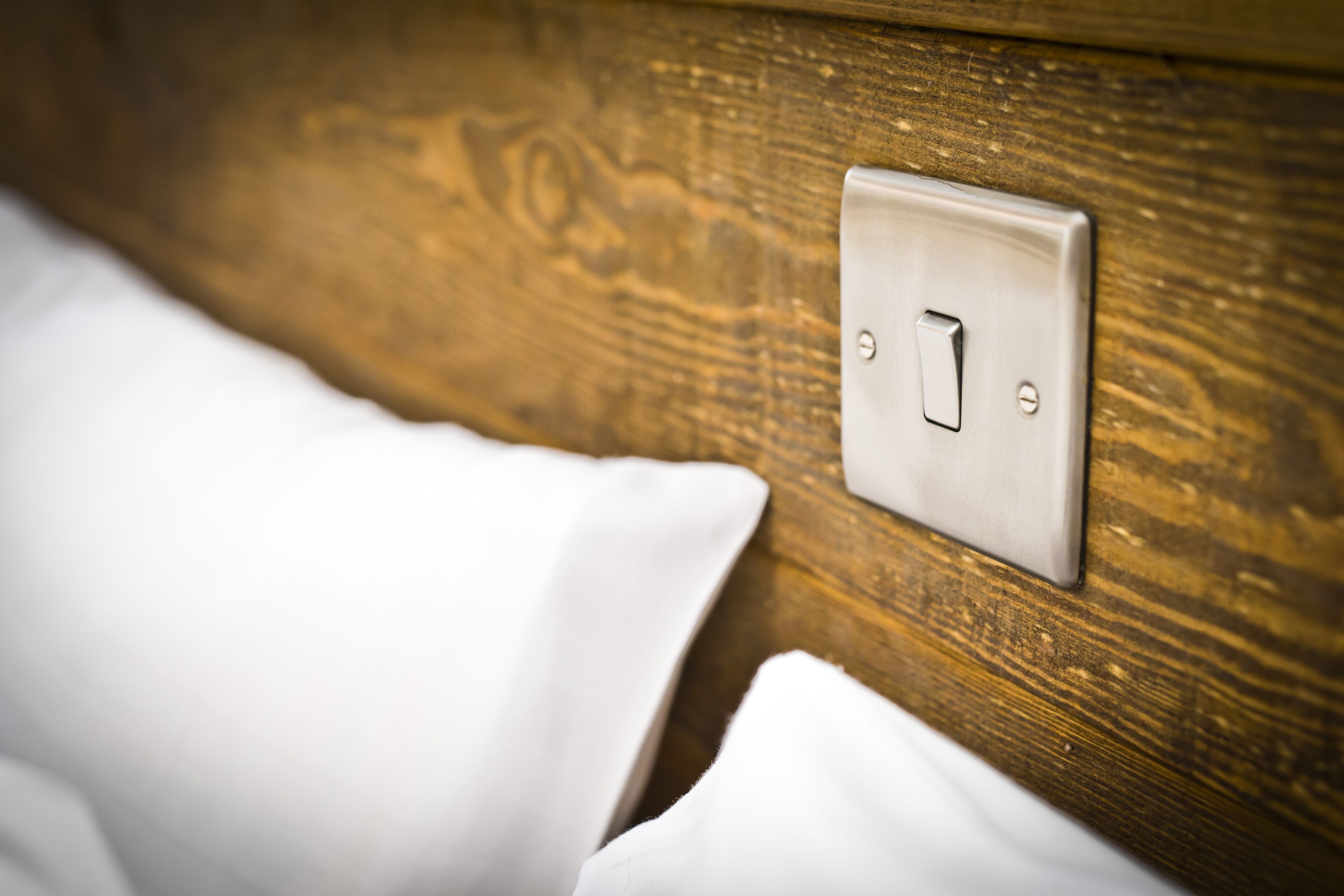 Detalhe de um interruptor de luz numa parede de madeira, um travesseiro branco e um lençol de cama branco.