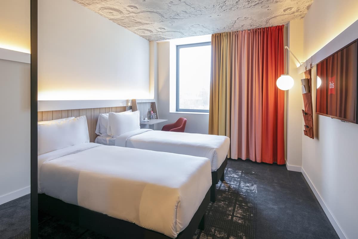 Quarto de hotel com duas camas, uma janela com cortinas, mesa de cabeceira, uma cadeira vermelha e uma luminária na parede.