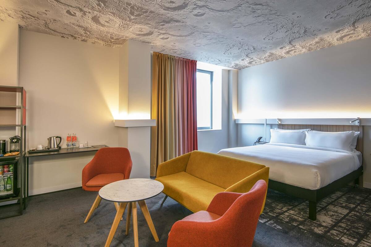 Quarto de hotel com cama, duas poltronas, uma mesinha redonda, um minibar e uma janela com cortinas.