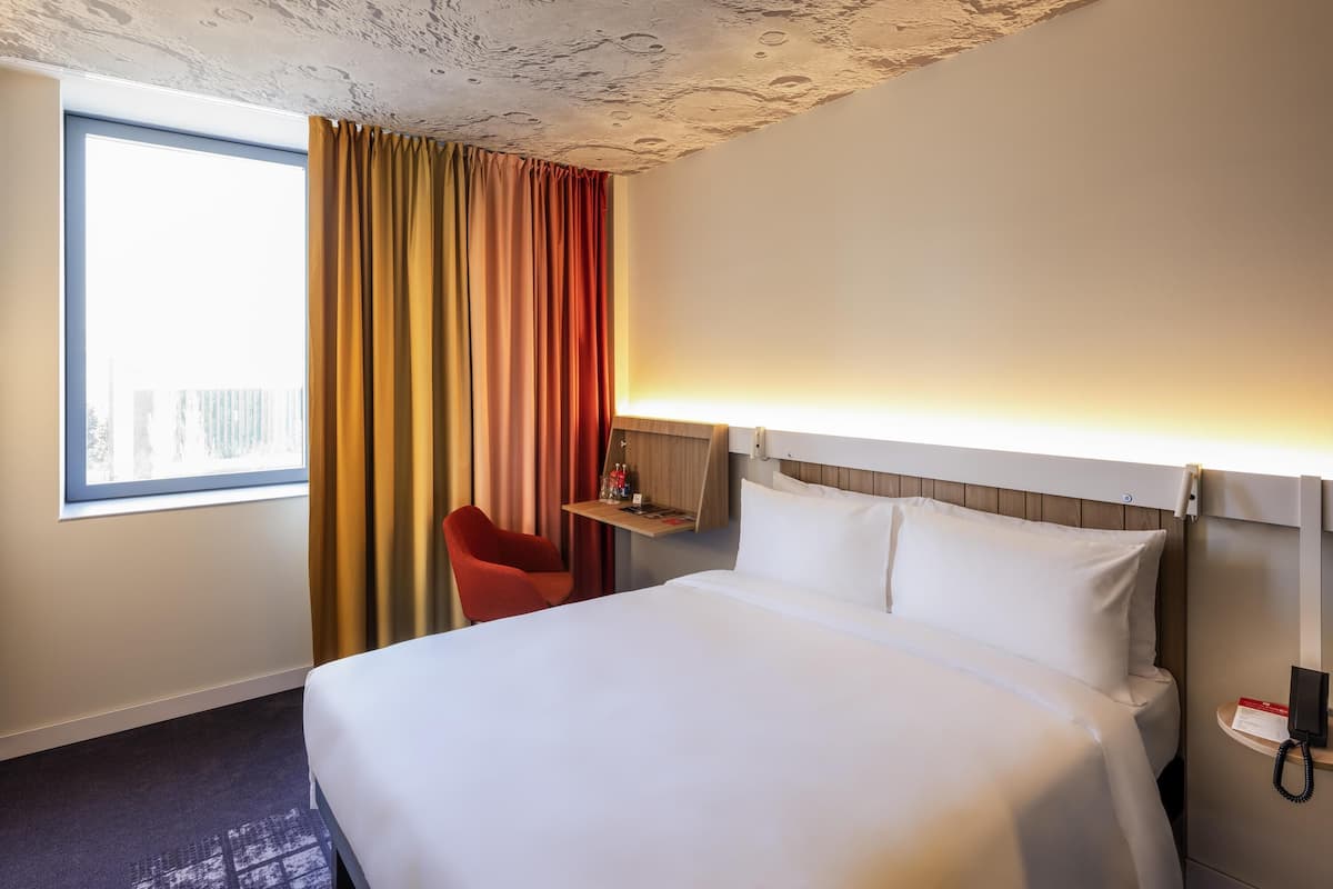 Quarto de hotel com cama, escrivaninha e cadeira, janela com cortinas e um design de teto único.