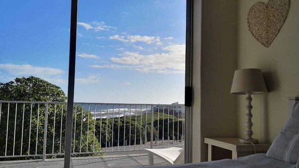 Uma varanda com grade de metal, uma cama com cabeceira branca, uma mesa de cabeceira com abajur e uma decoração de coração na parede.