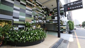 Entrada de hotel com fachada em azulejos, uma placa preta e um vaso com flores brancas.