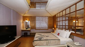 Quarto de hotel com cama, televisão, escrivaninha e uma janela grande.