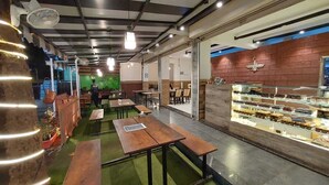 Um café com mesas e cadeiras de madeira, uma vitrine com produtos de panificação e uma parede de tijolos.