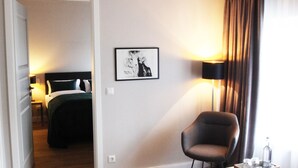 Quarto de hotel com cama, uma cadeira, mesa de cabeceira com abajur e um quadro na parede.