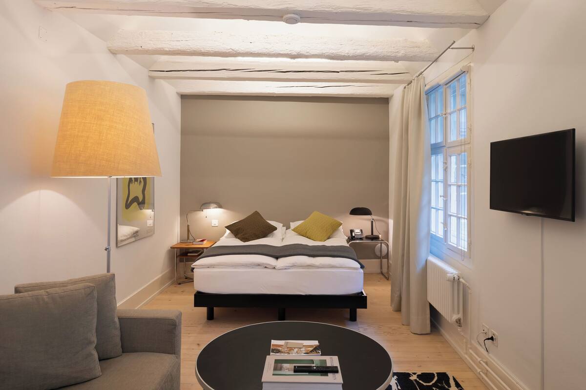 Quarto com cama, TV, luminária, sofá e janela.