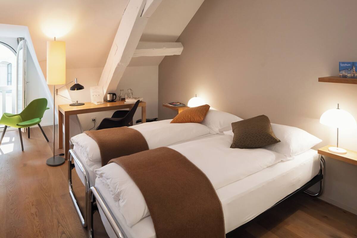 Quarto de hotel com duas camas, uma escrivaninha, uma cadeira, uma luminária, uma estante com livros e vista para o exterior.