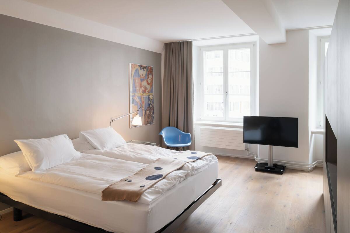 Quarto de hotel com uma cama, uma cadeira, uma televisão e uma janela com cortinas.