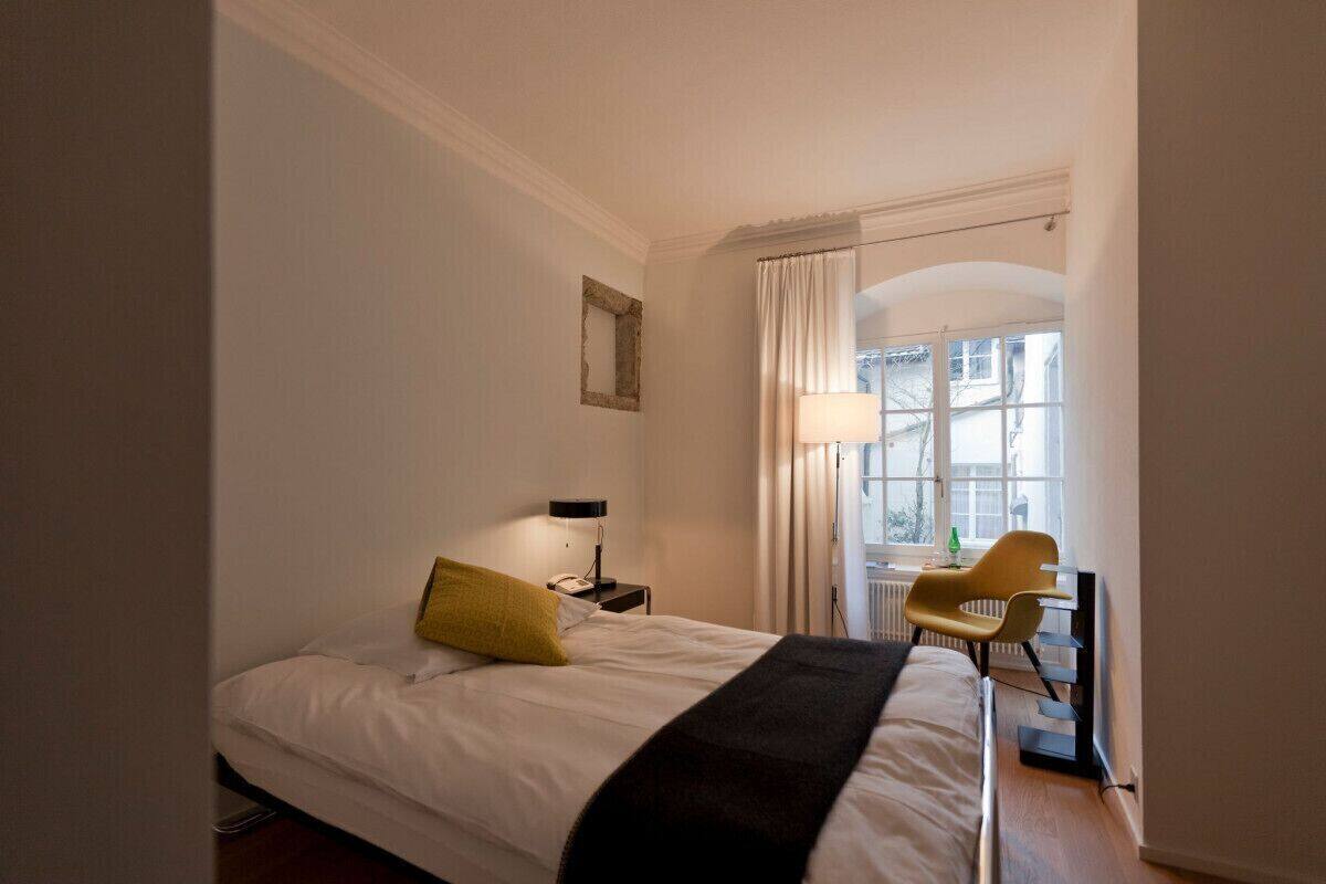 Quarto de hotel com cama, mesa de cabeceira, luminária, cadeira e janela com cortinas.