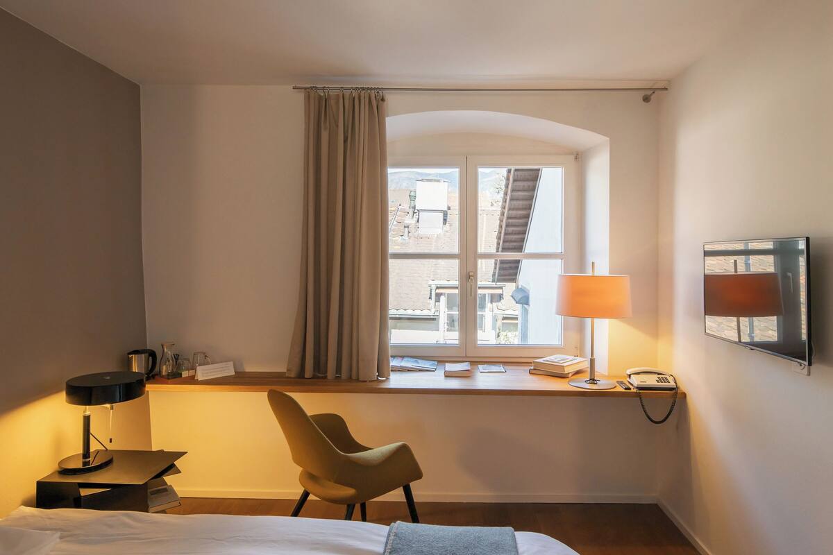 Quarto de hotel com uma cama, uma escrivaninha com luminária, uma cadeira, uma janela com cortinas, uma televisão e um telefone.