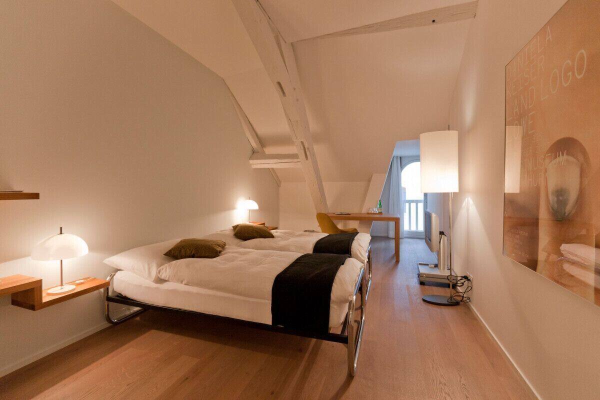 Quarto de hotel moderno com uma cama, abajures, uma mesinha de cabeceira de madeira, uma escrivaninha e um pôster grande na parede.