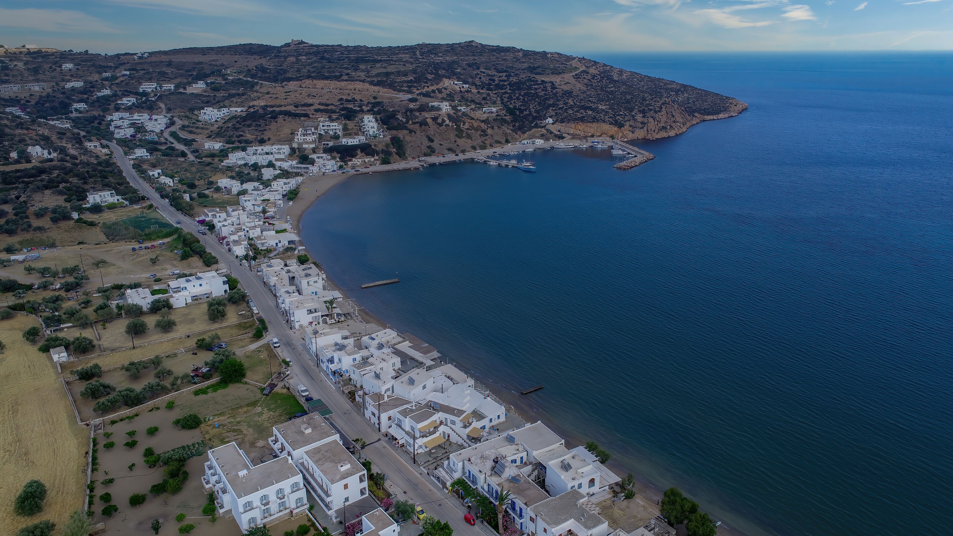 Vista aérea de uma cidade costeira com edifícios brancos, um porto de iates e um relevo montanhoso ao fundo.