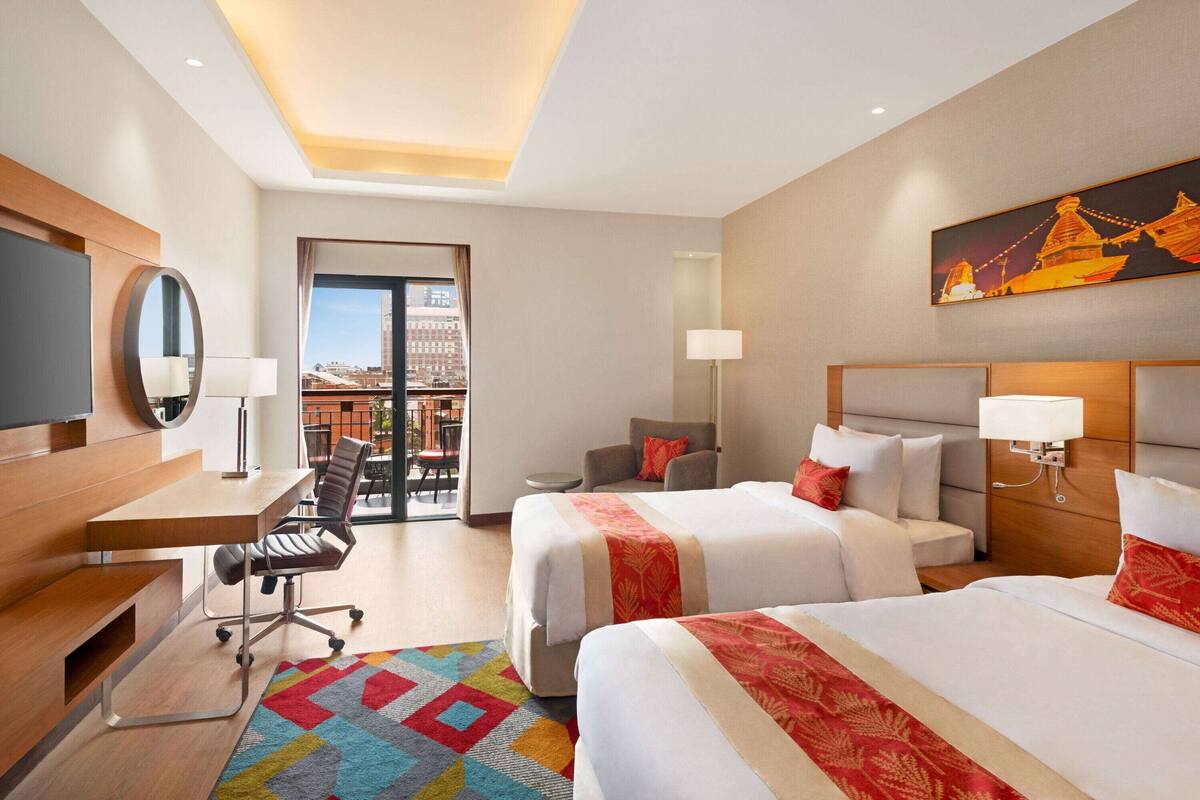 Quarto de hotel com duas camas, uma escrivaninha, uma cadeira e varanda com vista para a cidade.