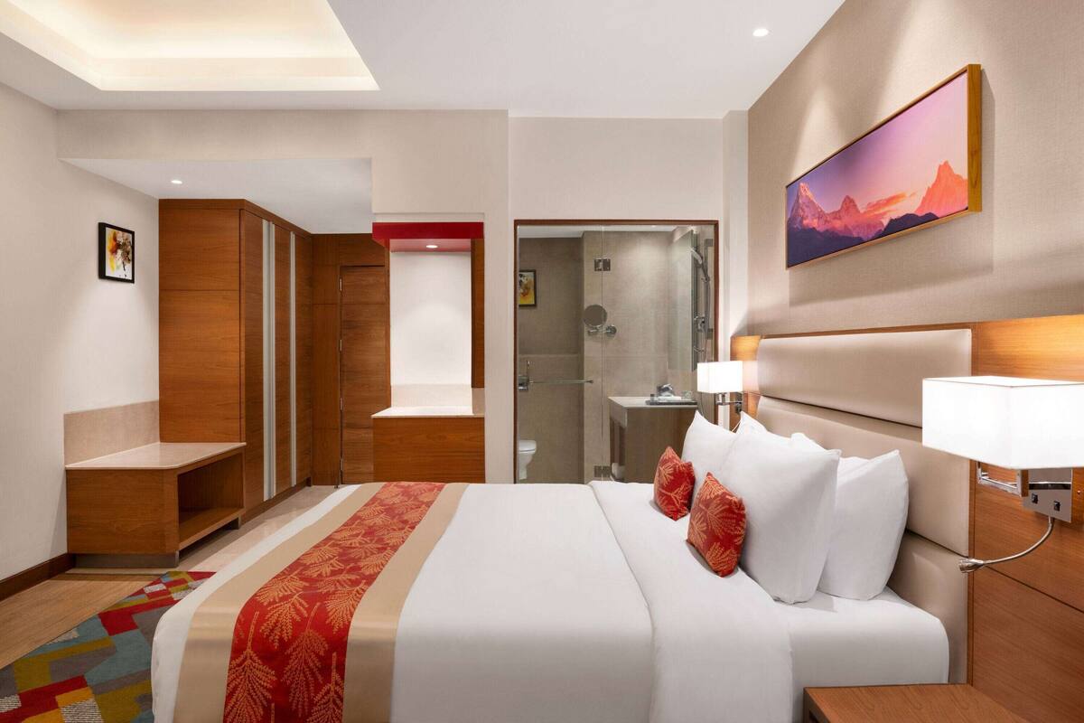Quarto de hotel com uma cama grande, uma mesa de cabeceira, uma área de chuveiro e televisão na parede.