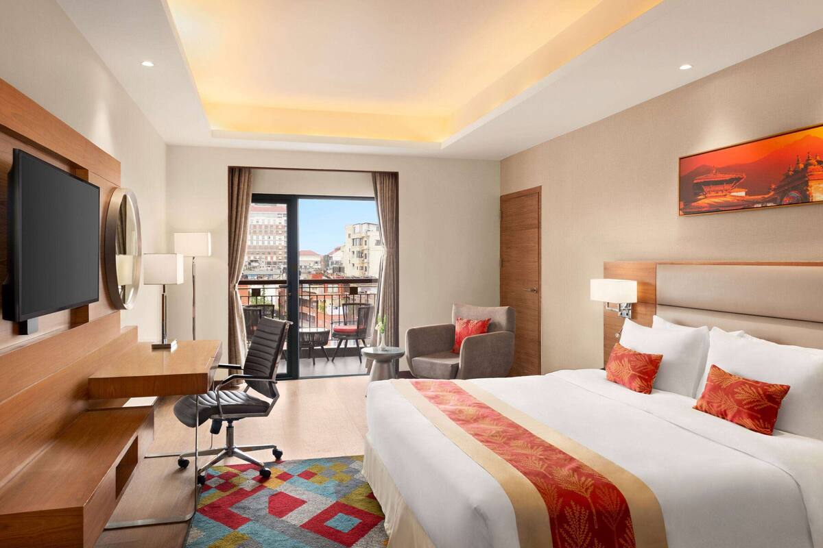 Quarto de hotel com uma cama grande, uma escrivaninha, uma cadeira, uma televisão e varanda com vista para edifícios.