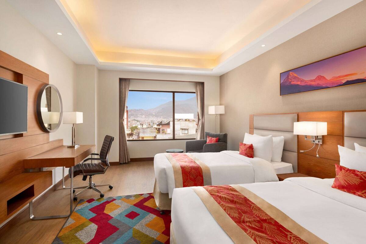 Quarto de hotel com duas camas, uma mesa, uma cadeira, televisão e uma janela grande com vista para as montanhas.