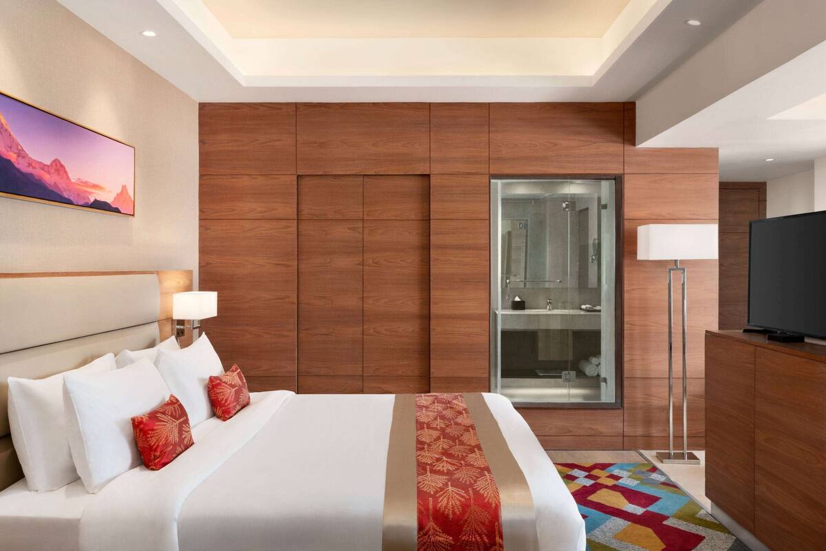 Quarto de hotel com cama, televisão, chuveiro e uma janela grande.