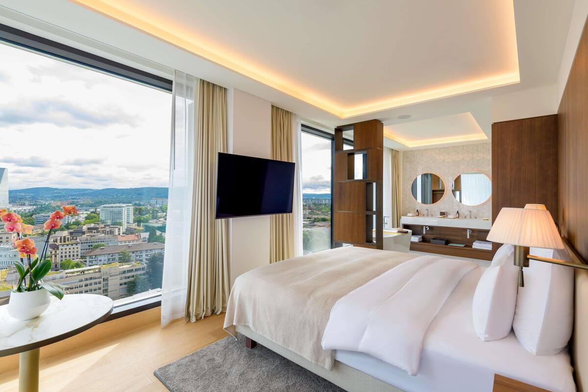 Quarto de hotel moderno com uma cama grande, televisão, banheiro com espelho e vista para a cidade.