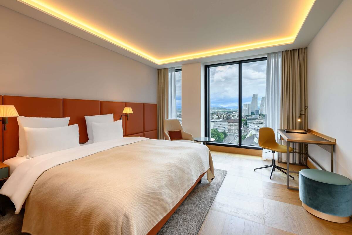 Quarto de hotel moderno com uma cama grande, uma escrivaninha, uma cadeira e vista para a cidade.