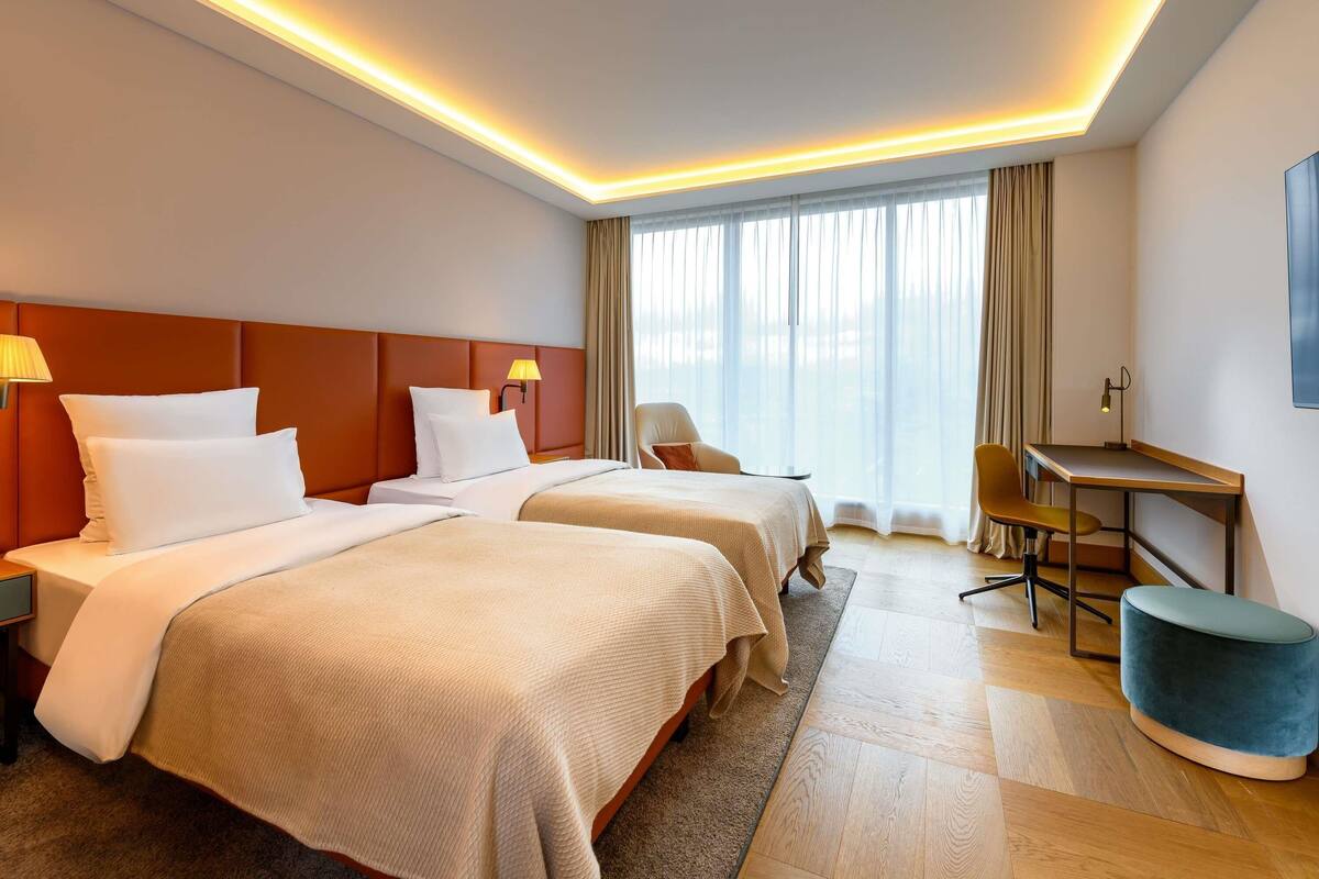 Quarto de hotel com duas camas, uma escrivaninha, uma cadeira e um pequeno banquinho.