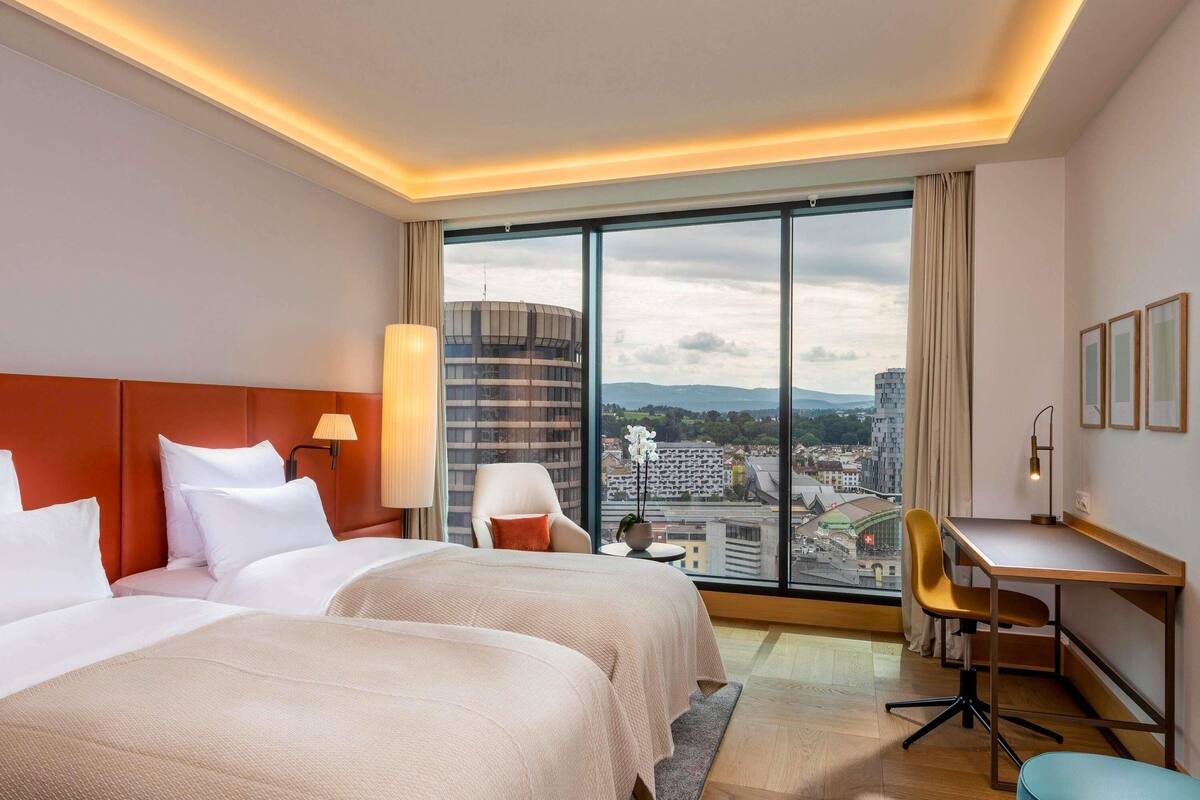 Quarto de hotel com duas camas, uma escrivaninha, uma cadeira, um abajur e uma janela grande com vista para a cidade.