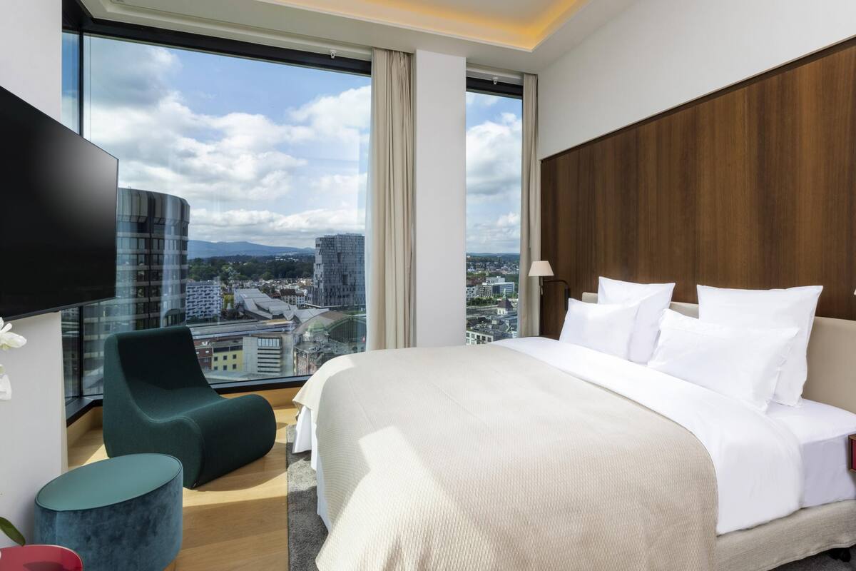 Quarto de hotel moderno com uma cama grande, vista para a cidade, televisão de tela plana e uma cadeira verde.
