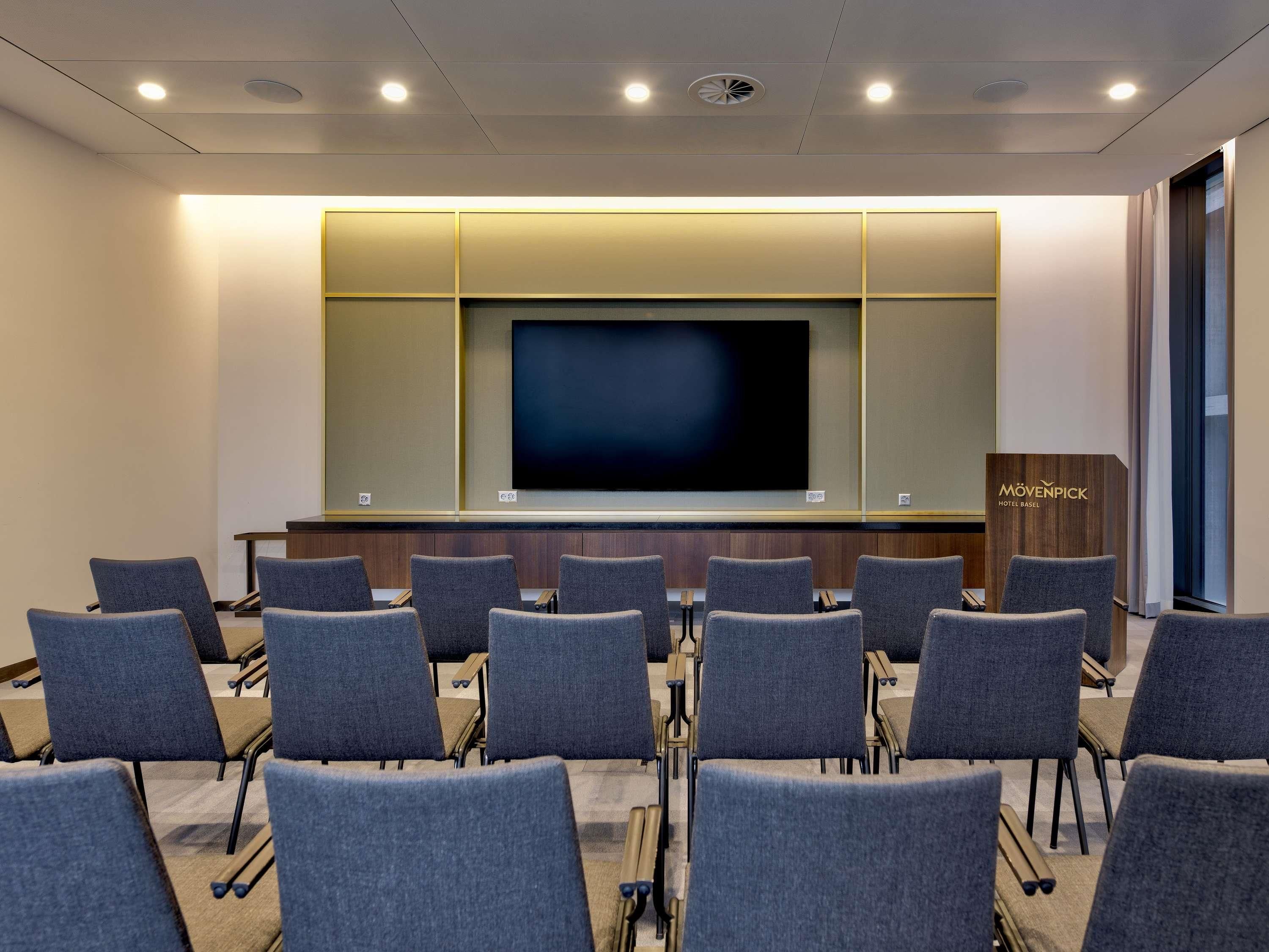 Uma sala de conferências com uma grande TV de tela plana, fileiras de cadeiras cinzas, um púlpito e um design moderno no teto.