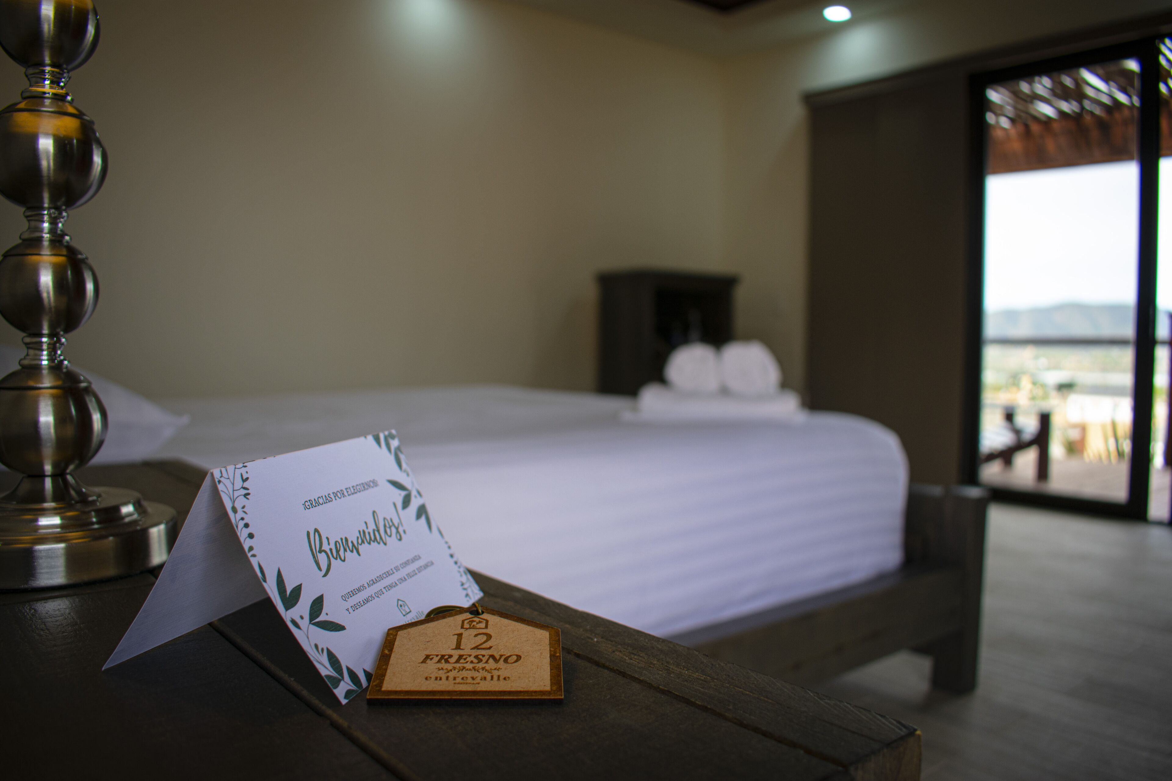 Quarto de hotel com uma cama bem arrumada, uma mesa de cabeceira de madeira com um cartão de boas-vindas, uma luminária e vista pela janela grande.