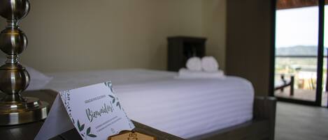 Quarto de hotel com uma cama bem arrumada, uma mesa de cabeceira de madeira com um cartão de boas-vindas, uma luminária e vista pela janela grande.
