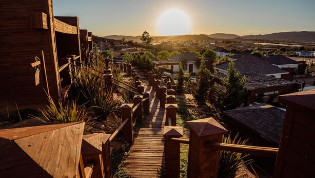 Uma escadaria de madeira leva a uma área residencial com casas e uma vista nítida do pôr do sol ao longe.