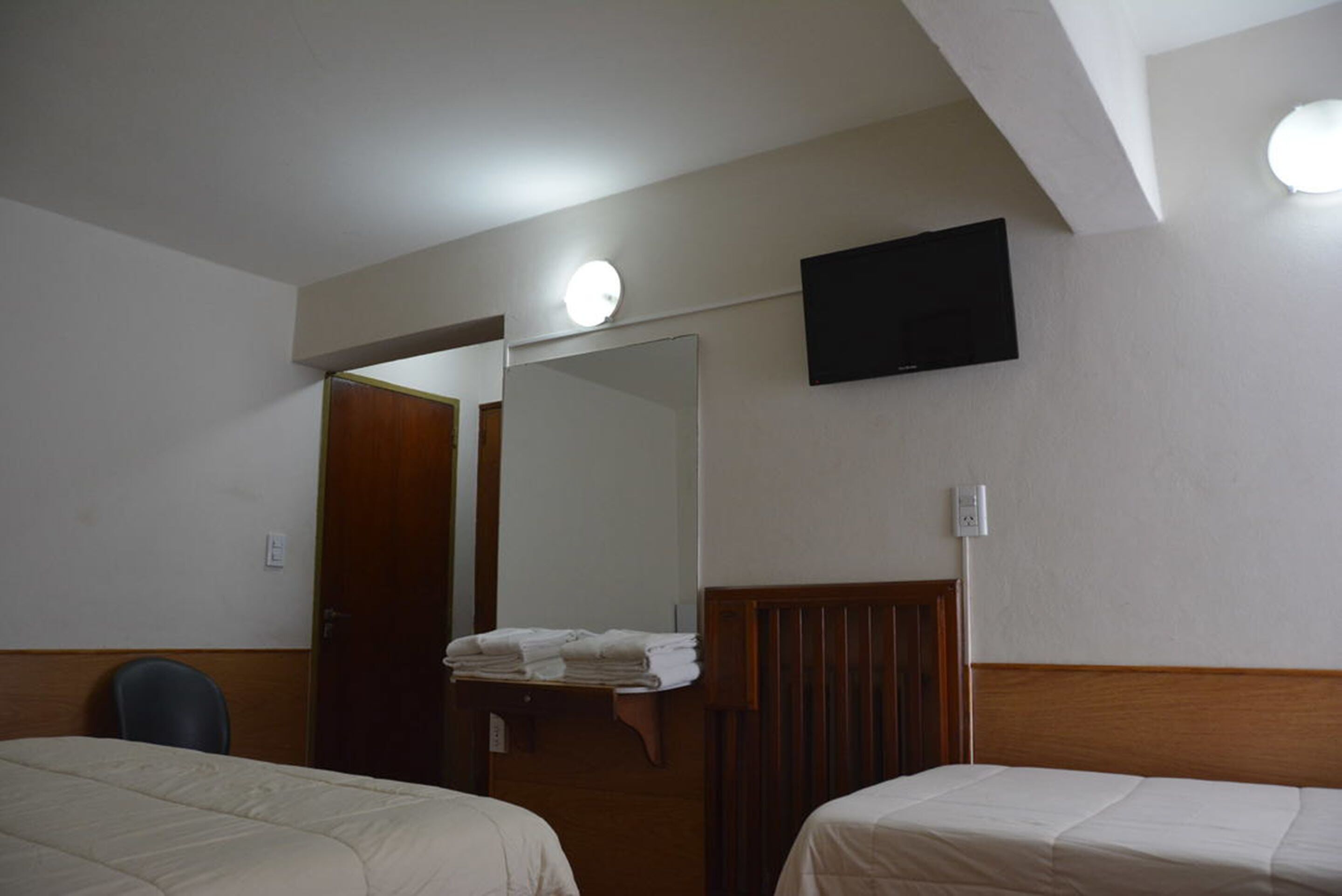 Quarto de hotel com duas camas, cabeceira de madeira, televisão de tela plana na parede e espelho sobre a porta.