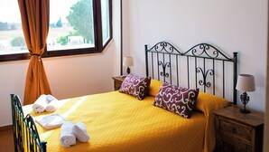 Cama bem arrumada com edredom amarelo e duas almofadas estampadas, janela com cortinas marrons e mesa de cabeceira com abajur.