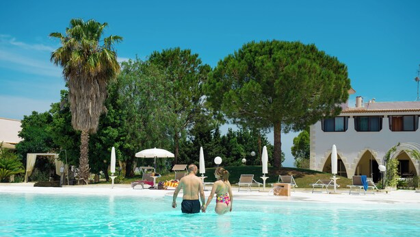 Piscina com espreguiçadeiras e guarda-sóis, rodeada por árvores e um edifício com janelas em arco.