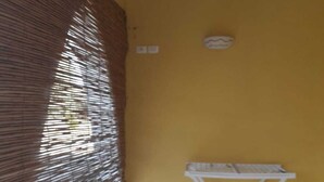 Quarto com parede amarela, janela com persianas marrons, tábua de passar branca e mesa com toalha de mesa florida.