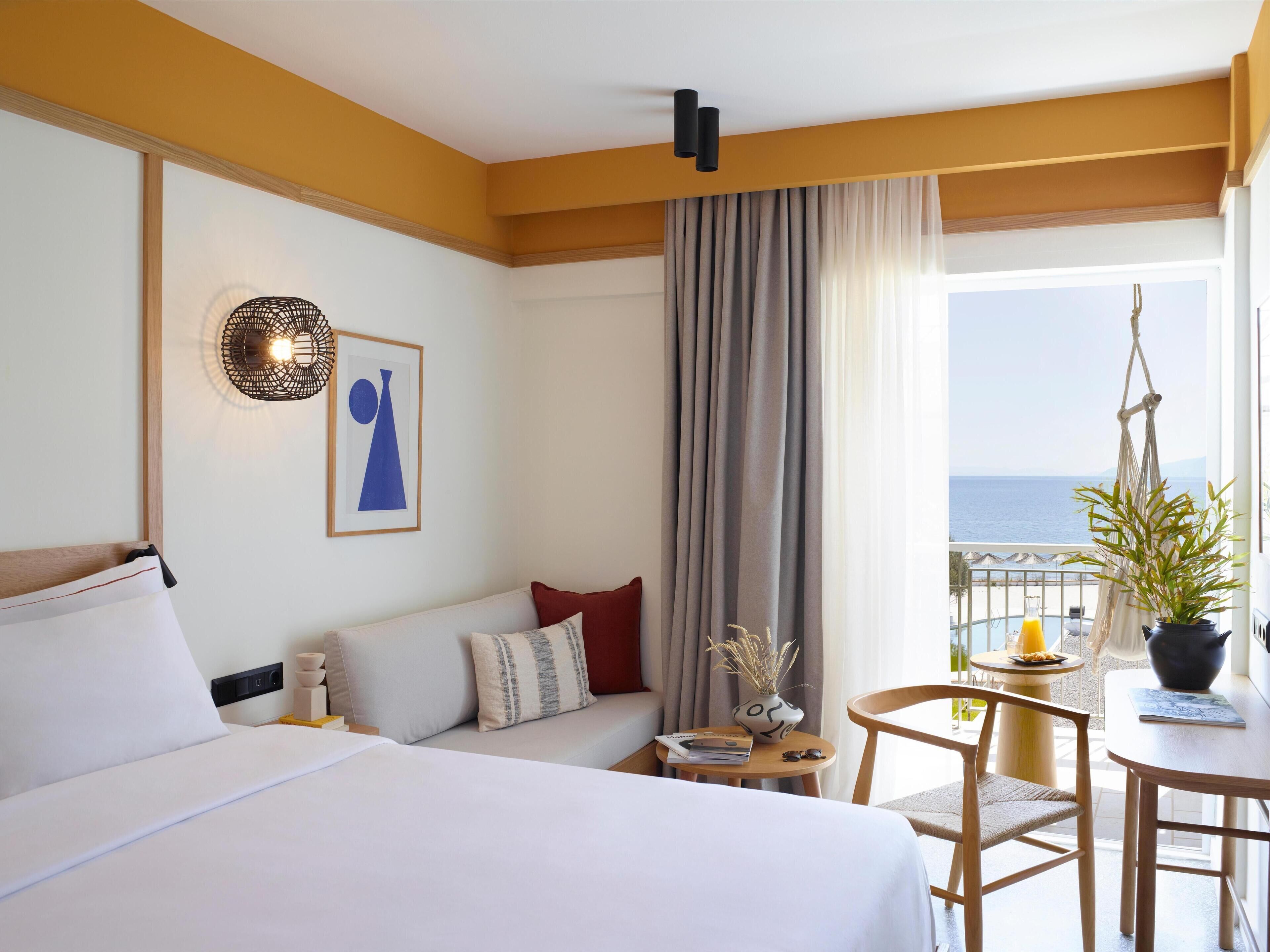 Quarto de hotel com uma cama, um sofá, uma mesinha com uma planta e vista para o mar.