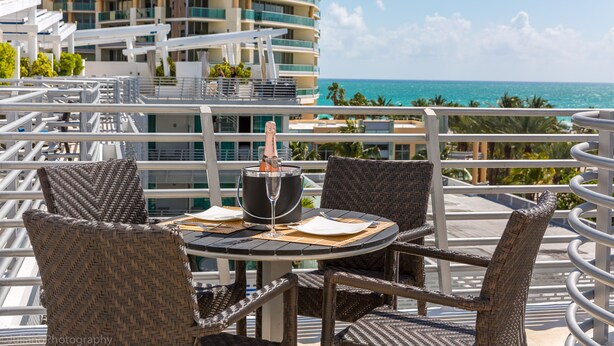 Varanda com cadeiras de vime e uma mesa, com vista para uma piscina e um edifício de frente para o oceano.