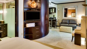 Quarto de hotel moderno com um armário curvo de madeira escura, uma TV de tela plana, um sofá e uma área de refeições com um espelho.