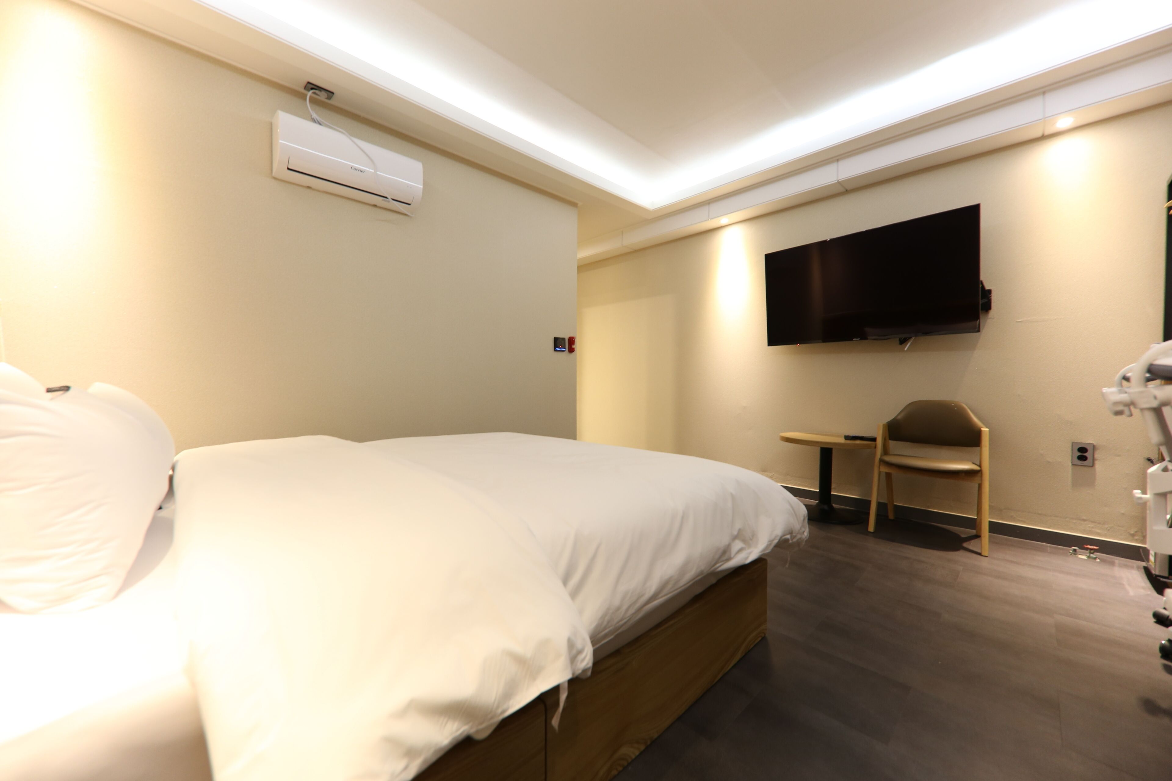Quarto de hotel com cama, poltrona, escrivaninha, televisão na parede e ar condicionado.