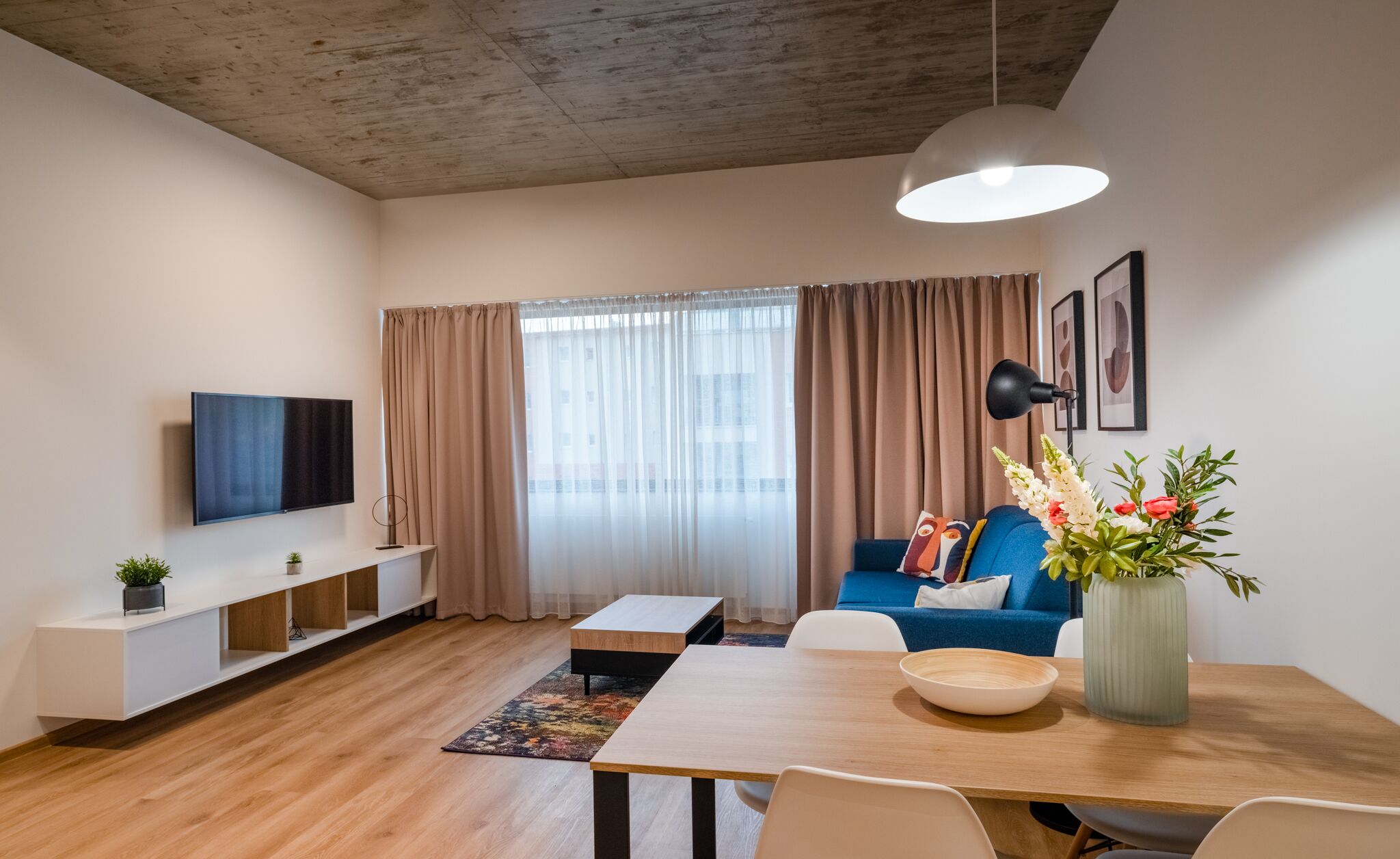 Uma sala de estar moderna com uma TV de tela plana, um sofá azul, uma mesa de jantar com cadeiras e uma janela grande com cortinas.