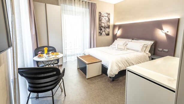 Quarto de hotel moderno com cama, mesa de cabeceira, cadeira e pequena área de trabalho.