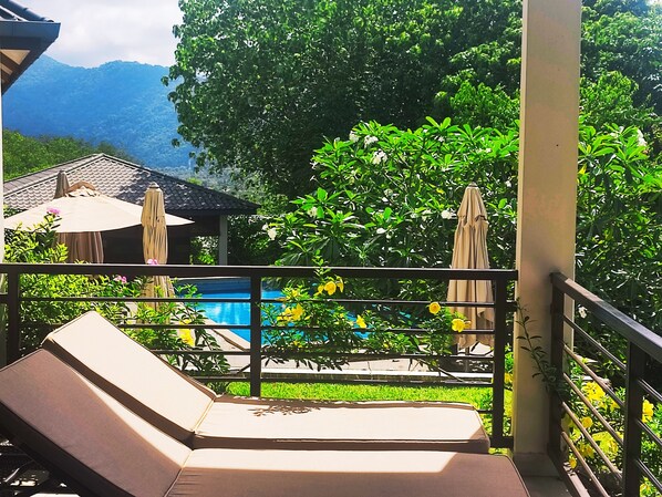 Uma varanda com vista para uma piscina, vegetação exuberante e montanhas ao fundo.