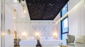 Quarto de hotel moderno com duas camas, uma mesa de trabalho e um sofá. O quarto possui teto estrelado e grandes janelas.