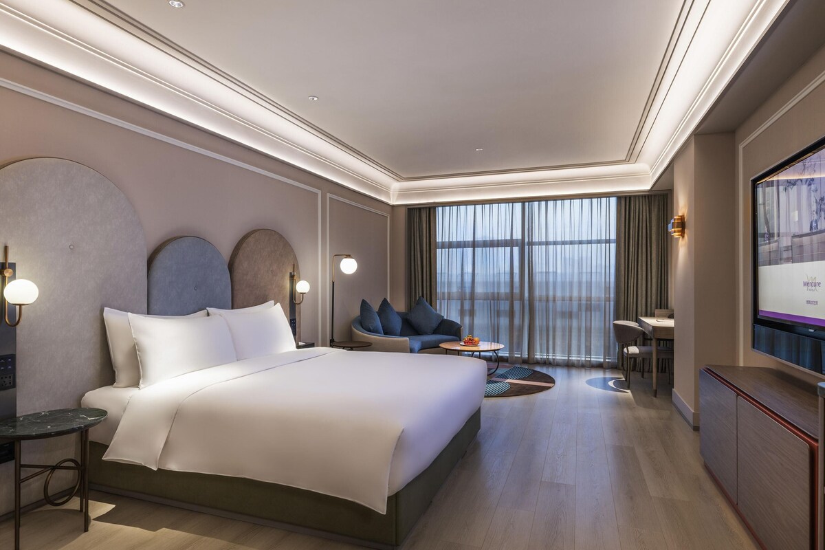 Quarto de hotel moderno com uma cama grande, área de estar com vista, escrivaninha e televisão.