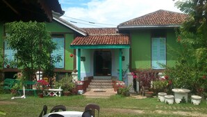 Uma casa de dois andares com telhado de telhas, paredes verdes e varanda frontal com degraus que levam à entrada.