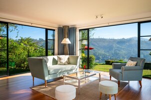 Uma sala de estar moderna com sofá, poltrona e mesa de centro, grandes janelas com vista para vegetação e colinas.