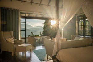 Quarto com uma cama grande, uma cadeira, uma mesinha e vista para as montanhas pela janela.