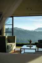 Quarto com uma grande janela que oferece vista para uma paisagem montanhosa. Há duas cadeiras e uma mesinha com um vaso.