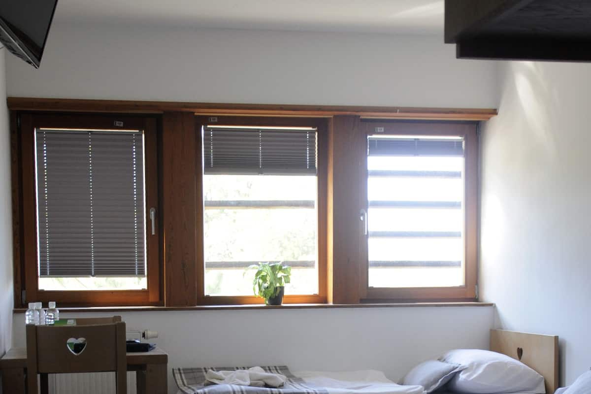Quarto com duas camas, uma escrivaninha e uma cadeira. Há janelas com persianas, uma planta no parapeito e uma televisão fixada na parede.