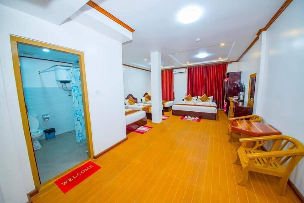 Quarto de hotel com piso de madeira, cama com cortina vermelha, área de estar com mesa e cadeiras, e banheiro visível por uma porta de vidro.