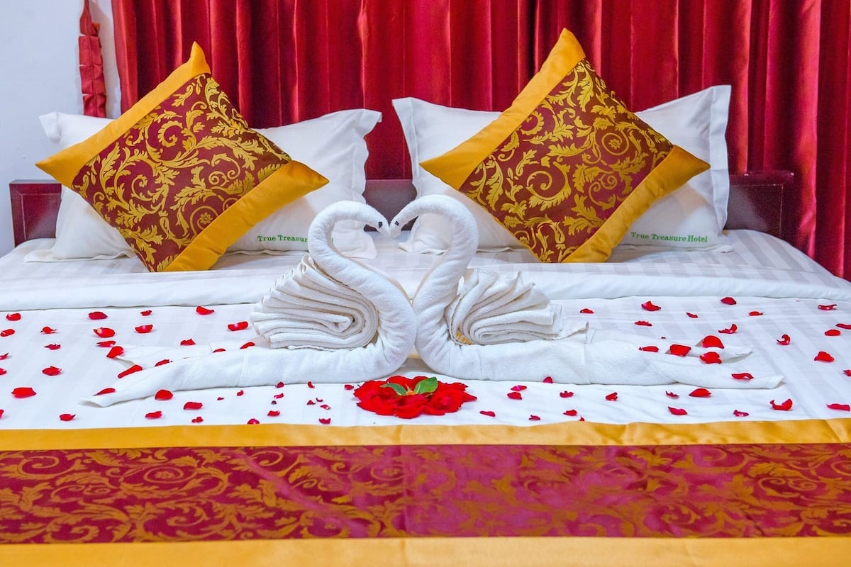 Cama com roupa de cama branca, duas almofadas decorativas com desenhos dourados e arranjo de toalhas em formato de coração.